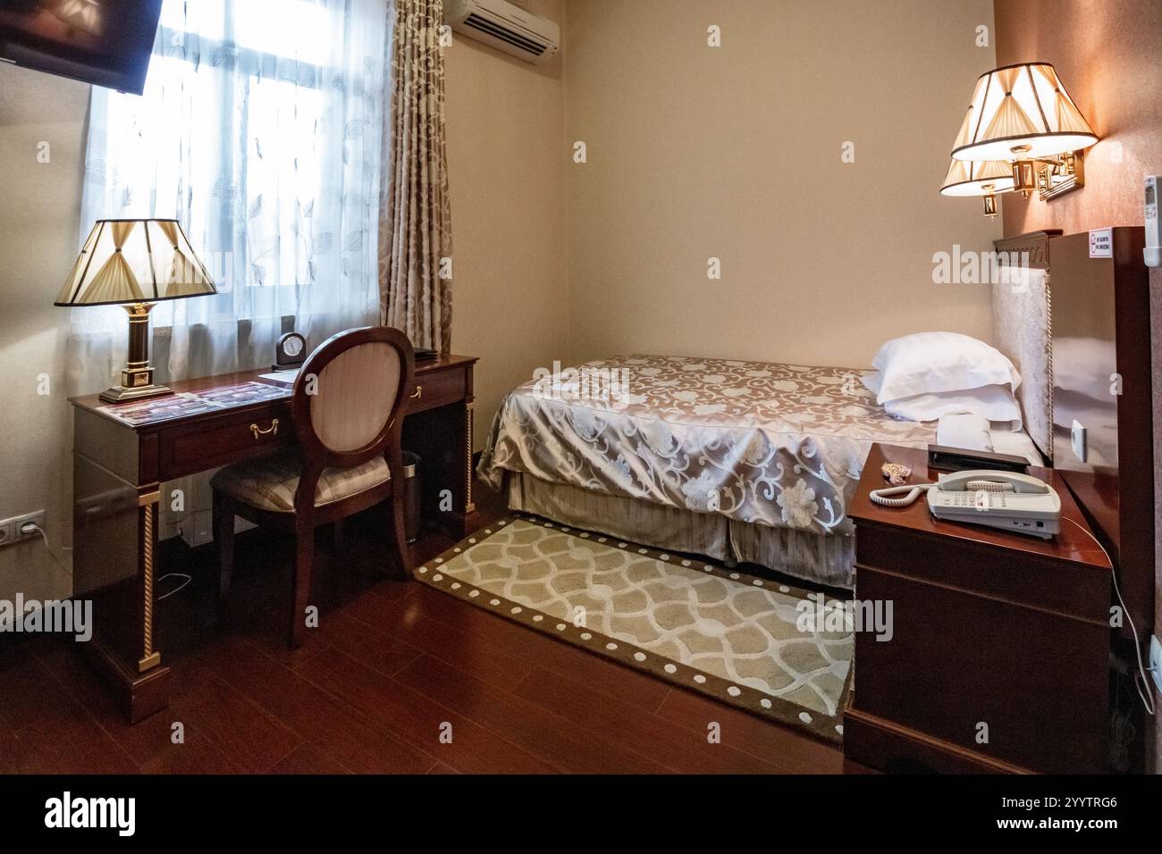 A small bedroom with a desk and a chair. The desk has a lamp and a ...