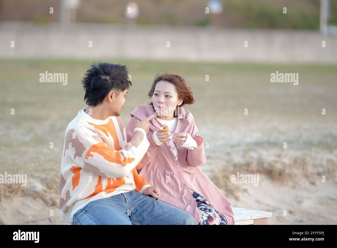 A couple in their 20s are happily eating crepes on a cold beach bench ...