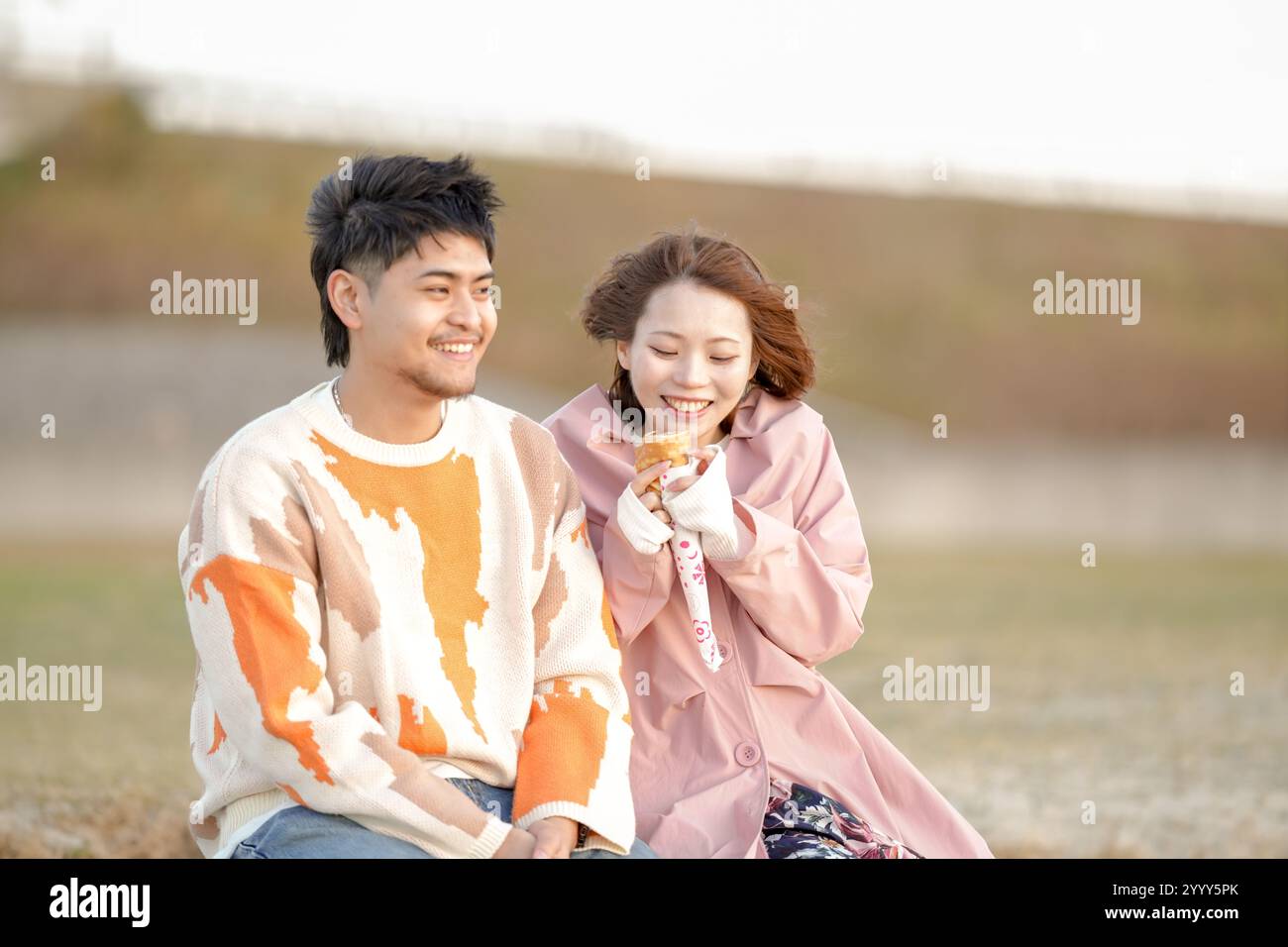 A couple in their 20s are happily eating crepes on a cold beach bench ...