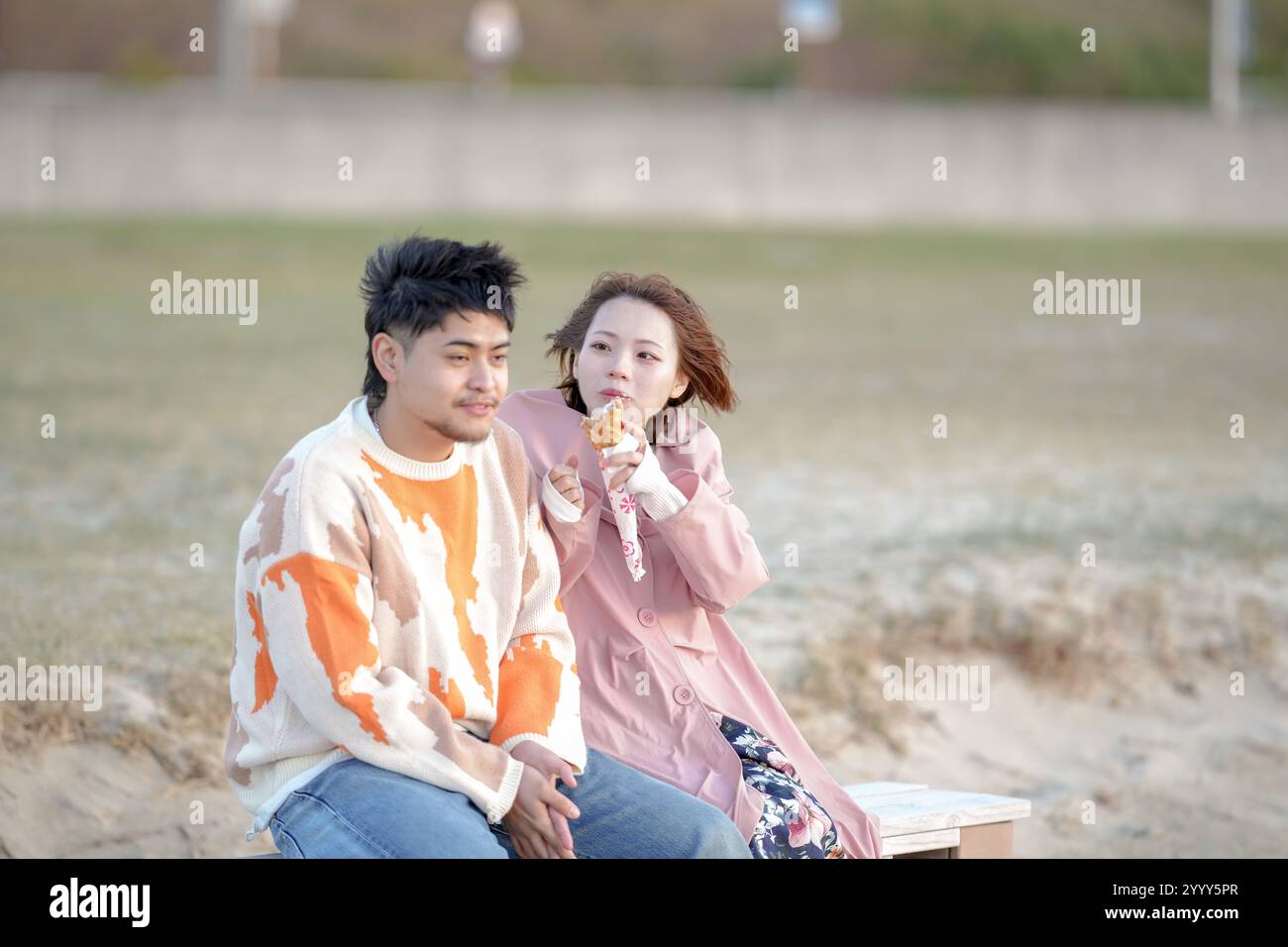 A couple in their 20s are happily eating crepes on a cold beach bench ...