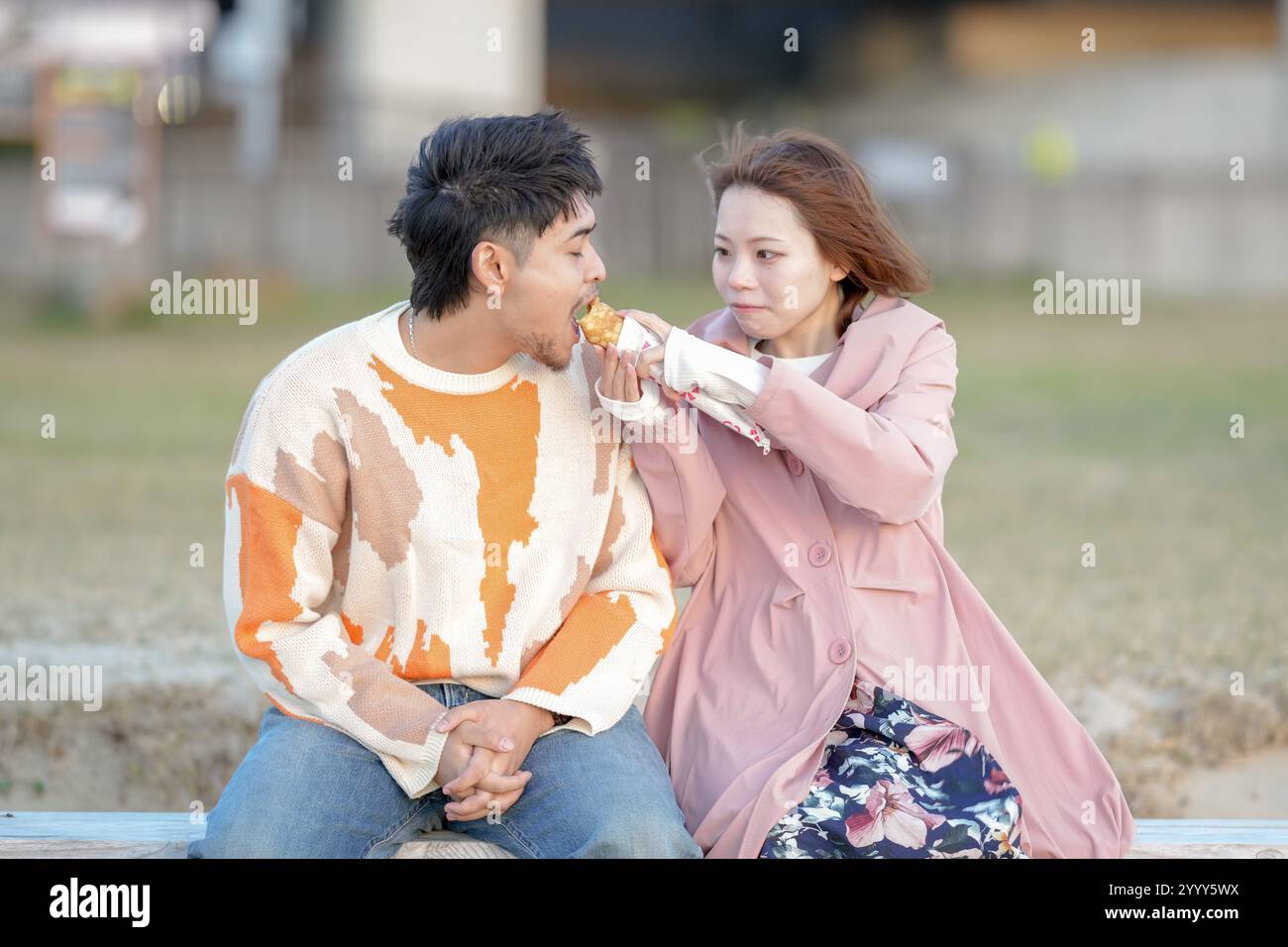 A couple in their 20s are happily eating crepes on a cold beach bench ...