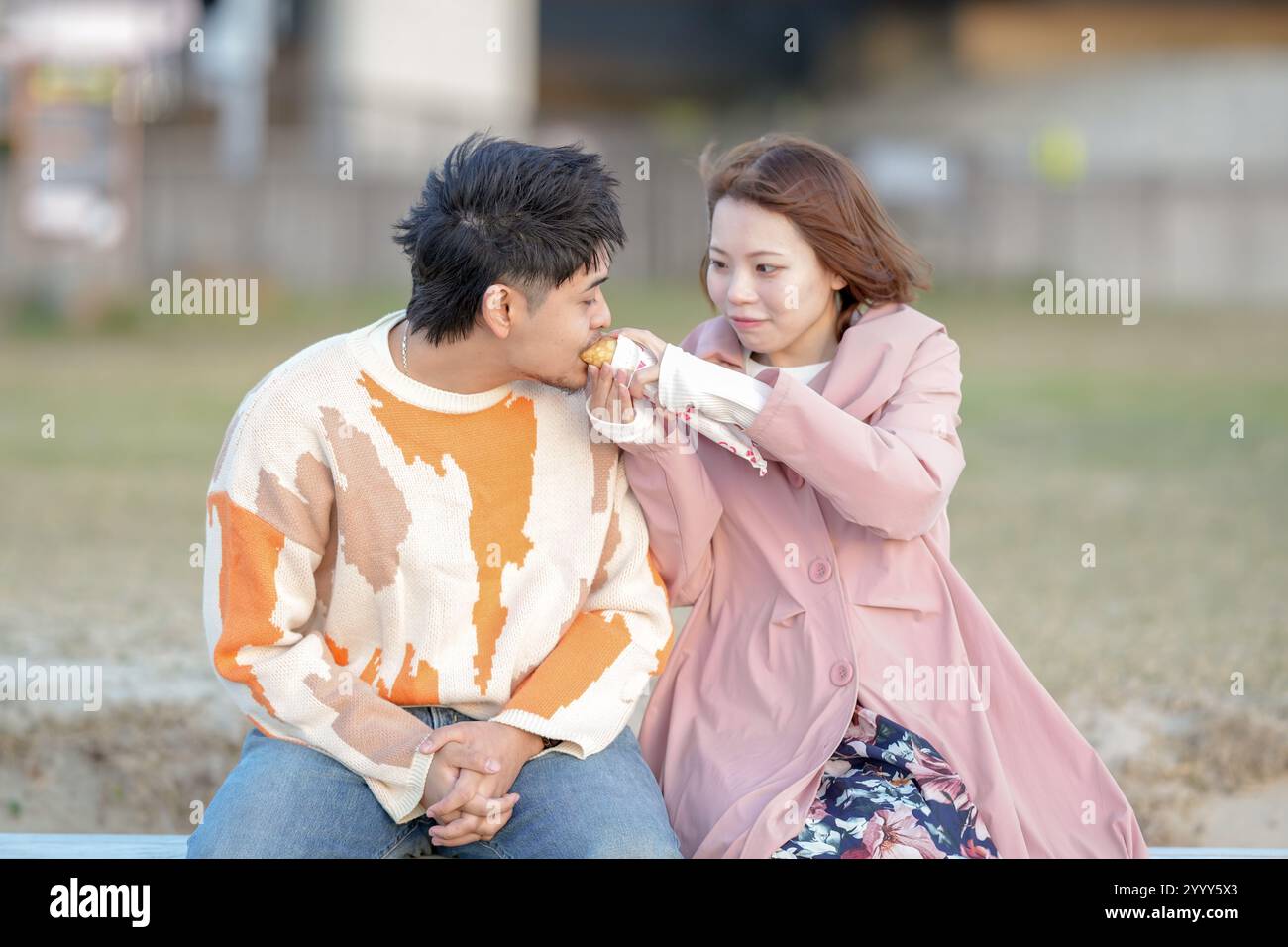 A couple in their 20s are happily eating crepes on a cold beach bench ...