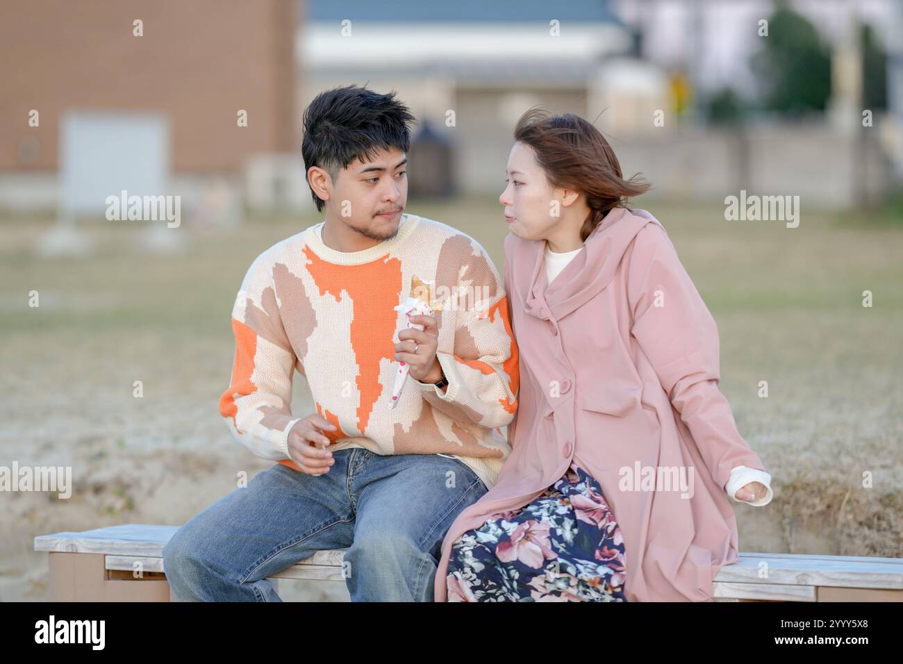 A couple in their 20s are happily eating crepes on a cold beach bench ...