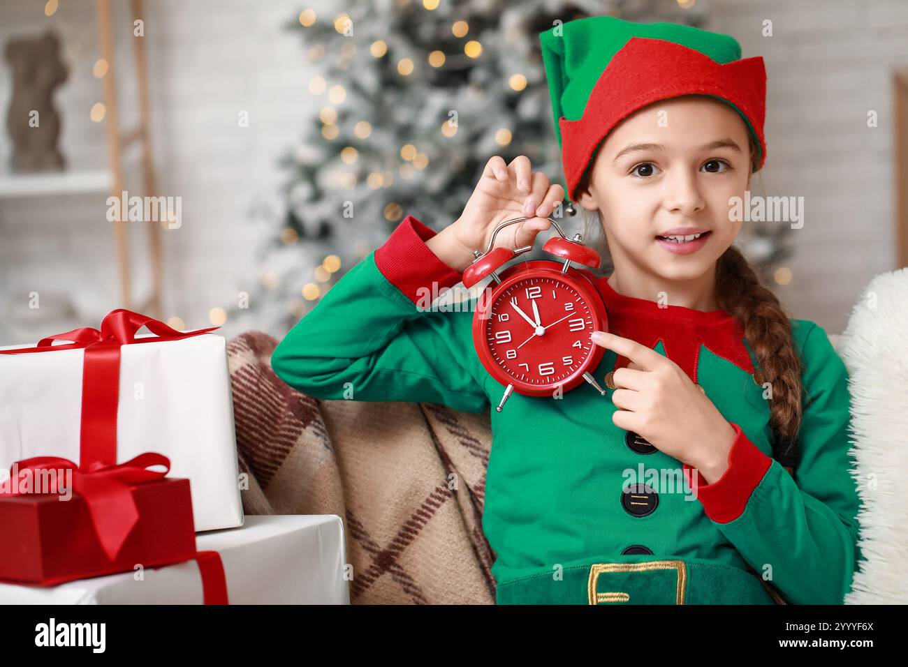 Cute little girl dressed as elf pointing at alarm clock in room on ...