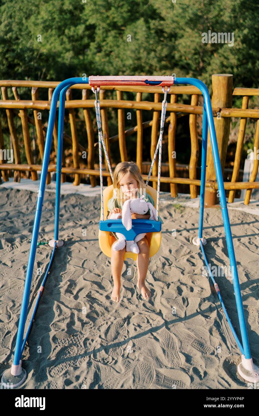 Little smiling girl swinging on a swing hugging a pink bunny at the ...