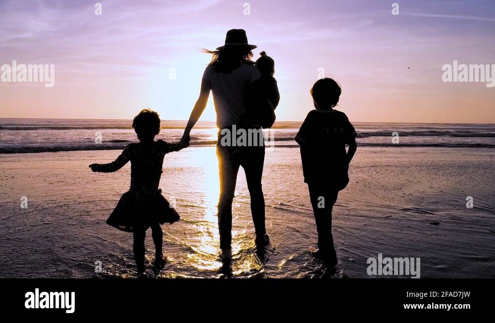 A mother holds hands and walks with her three kids at the beach in slow ...