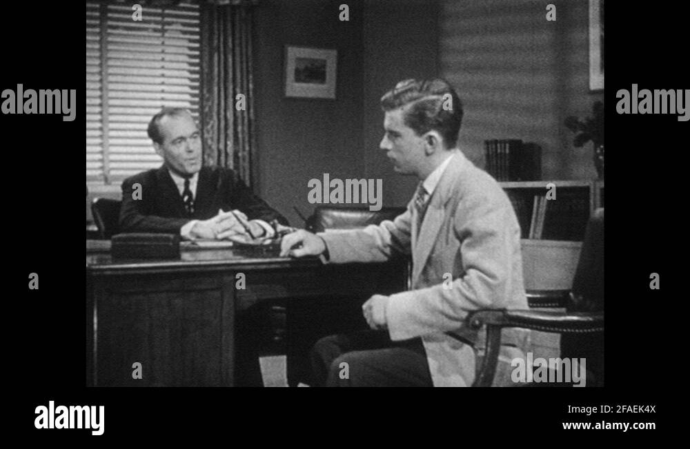 1940s: Two men sit across from each other at a desk, talking. One man ...