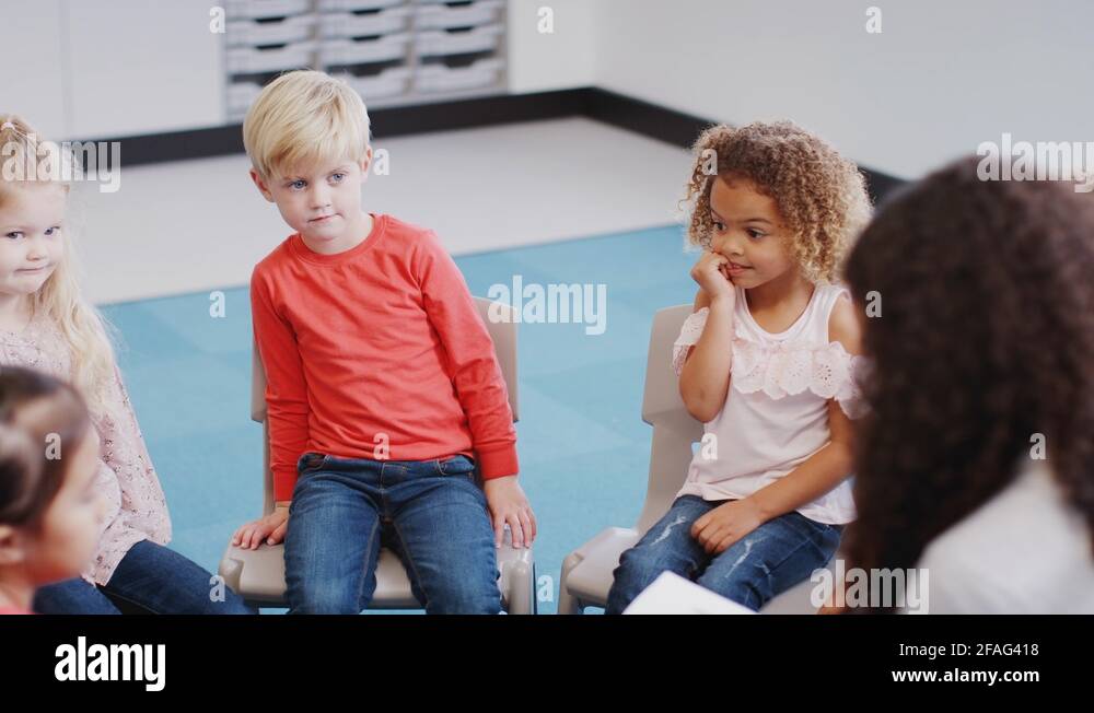 Infant school kids sitting on chairs in classroom listening their ...