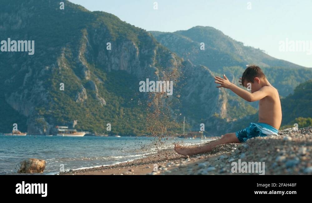 Boy sitting on beach and throwing sand and pebbles into sea Stock Video ...