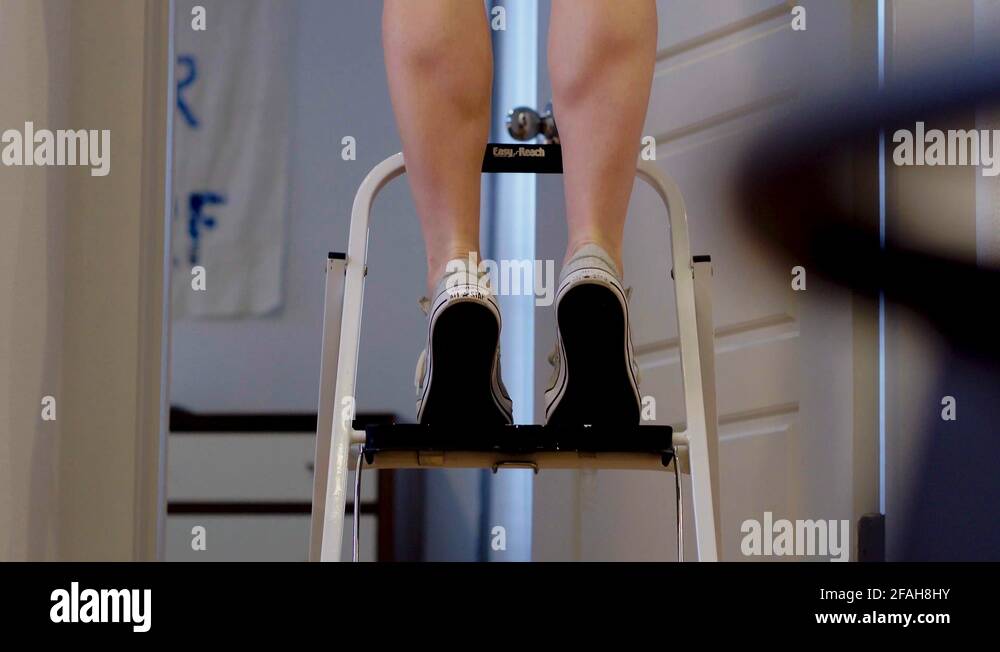 A woman on her toes on the top step of a step ladder reaching high up ...