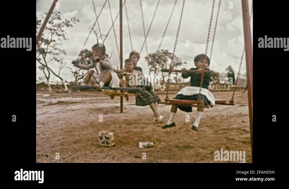 1960s: Children play on swing set in playground. Cars and people in ...