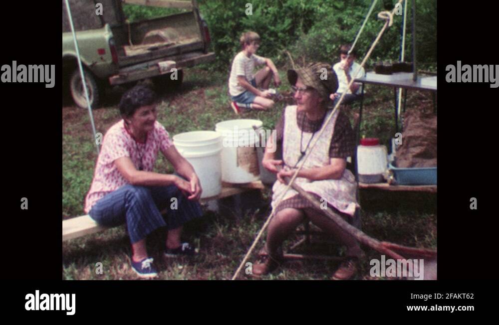 1970s: Two women sit on benches in grass and talk, one woman stirs stew ...