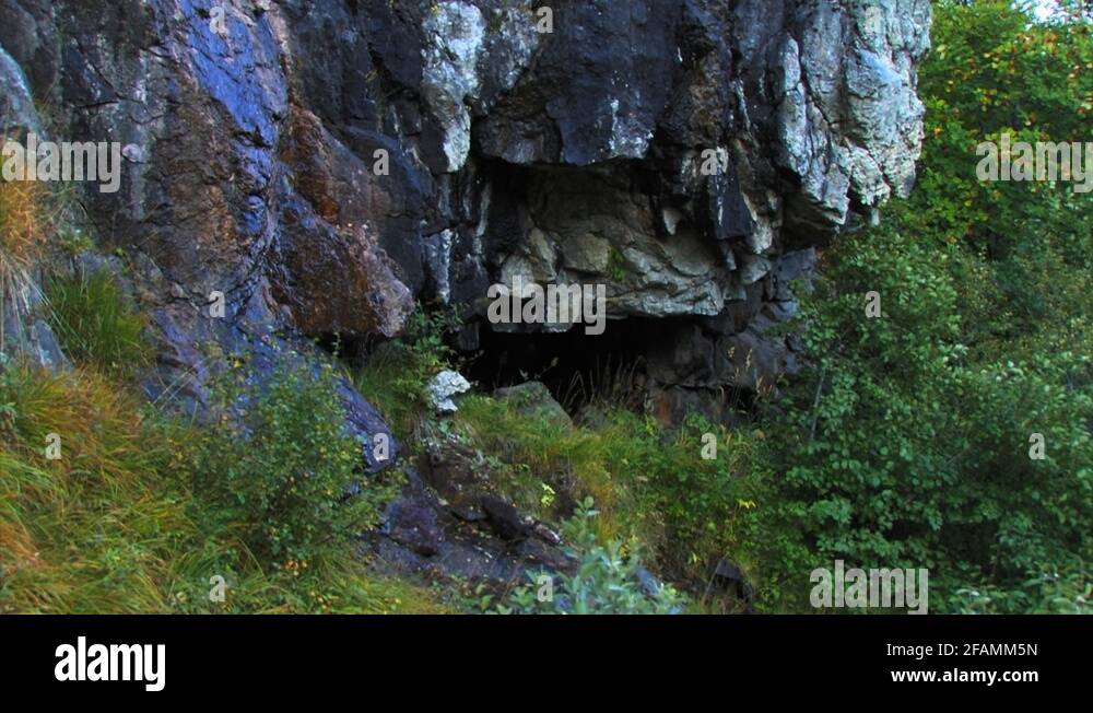 Zoom in on a jagged rock face and a cave entrance at the base. Trees ...