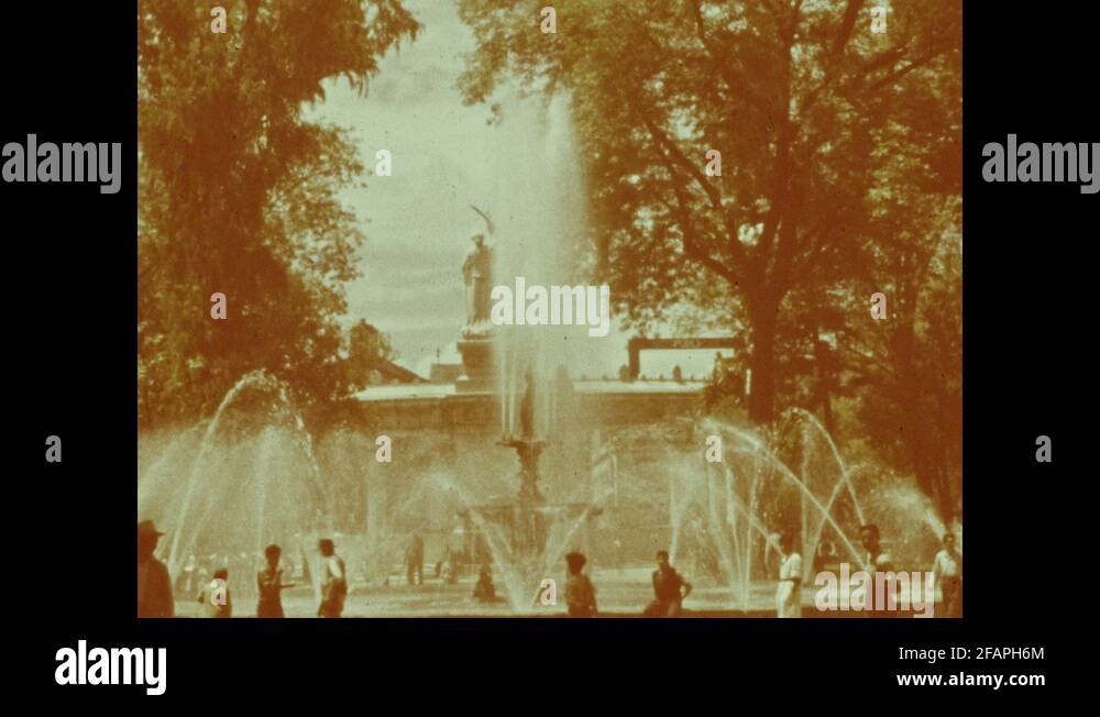 1950s: Busy park, tall trees, people walking, fountain, monument with ...