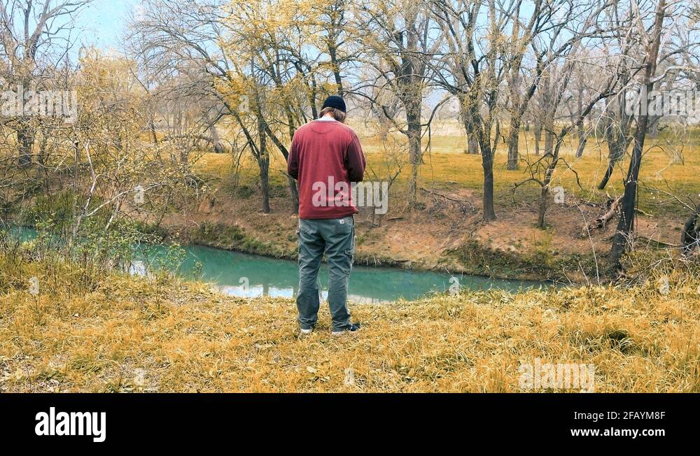 Grief stricken man stands alone at river in woods, slow pulling phone ...