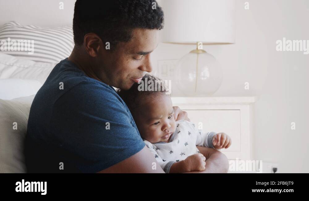 Millennial African American dad rocking in an armchair holding his baby ...