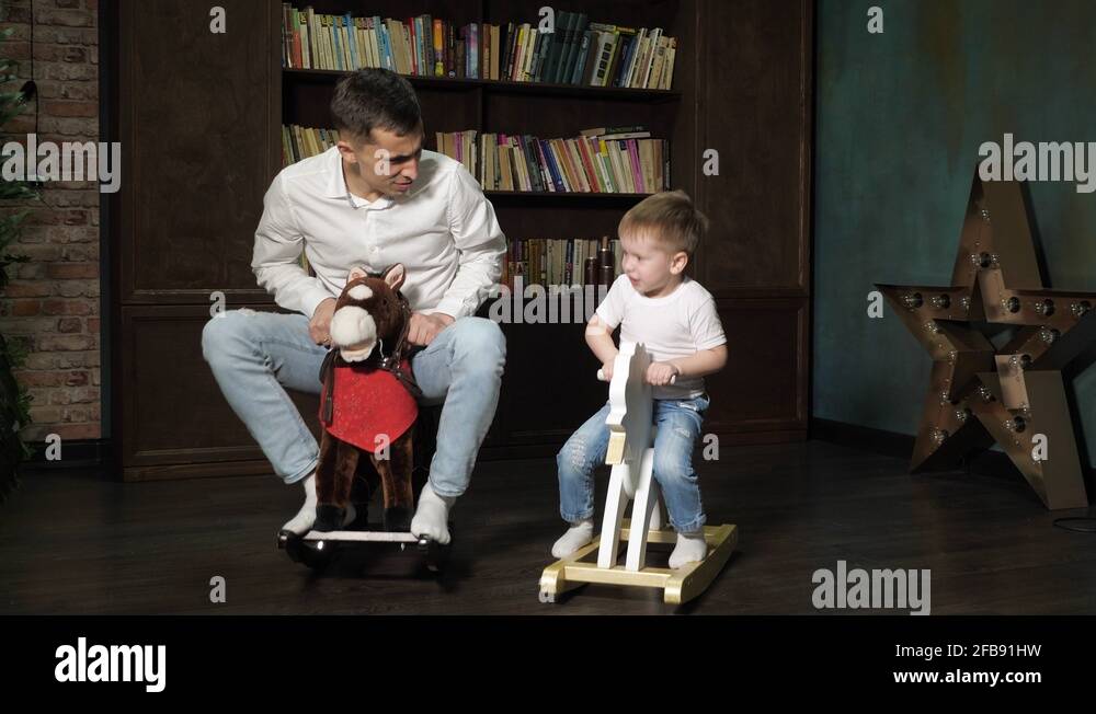 Dad and son are playing together riding on toy horses rocking chair ...