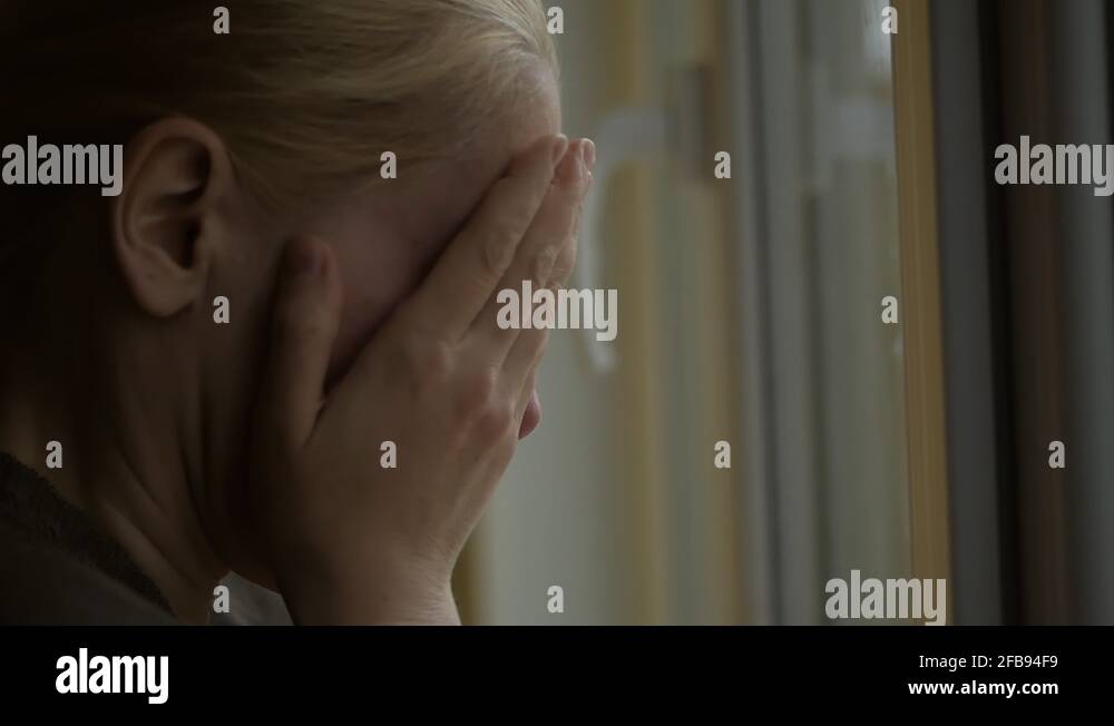 Woman standing at the window in the room crying, covering his face with ...