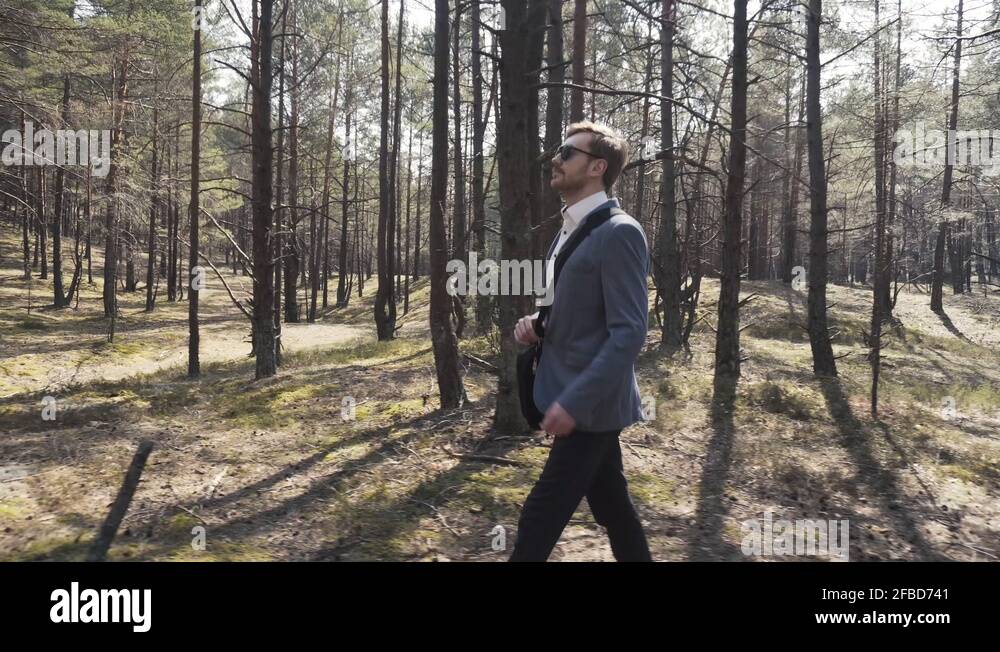 Young man dressed in suit walking trough sunny pine tree forest ...