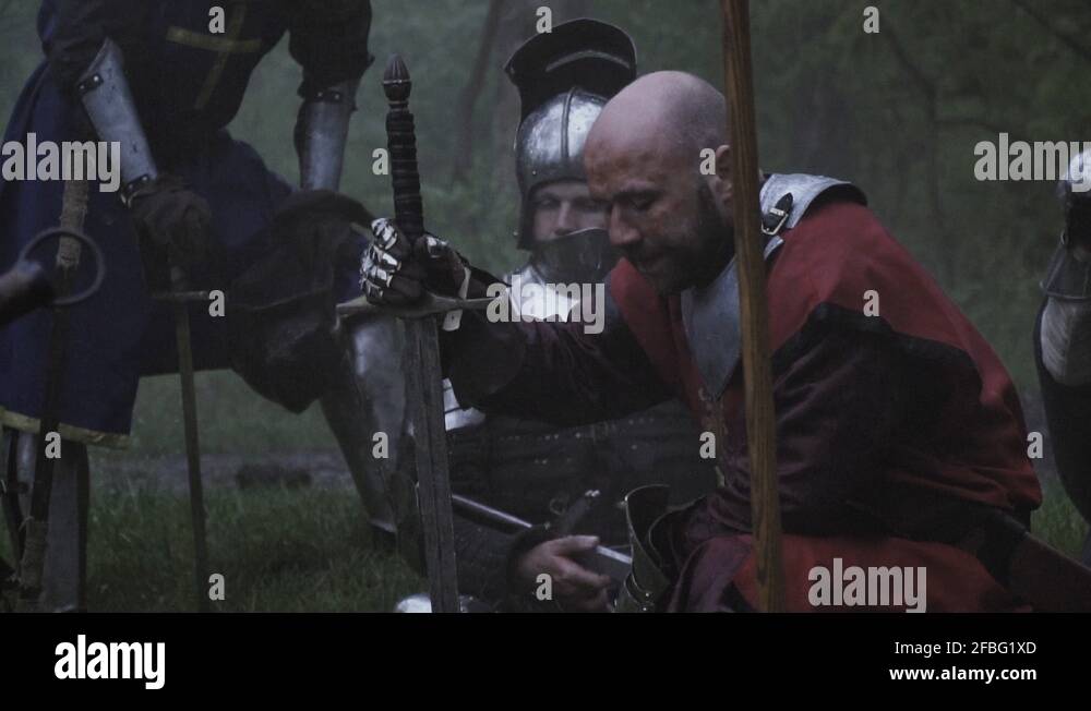 Knight in red armor is kneeling with his sword in hand, under rain ...