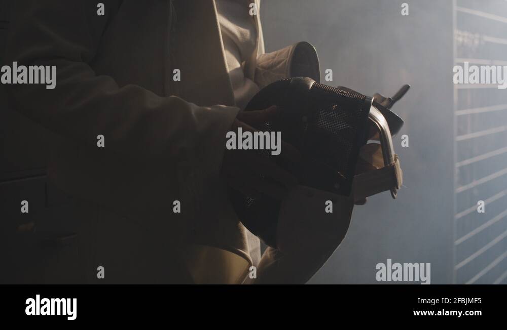 Man in fencing uniform checks out his fencing helmet in a moody gym ...
