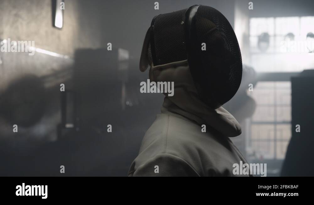 A fencer takes off his fencing mask in a dark and foggy locker room ...