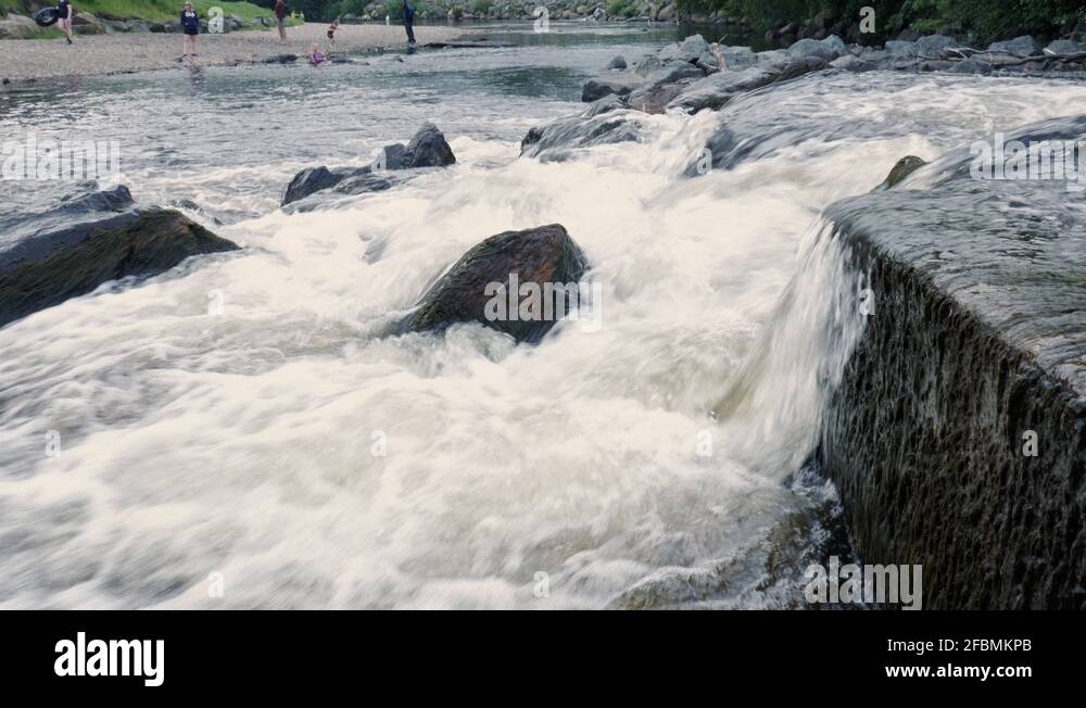 A tiny low waterfall near a metal bridge; people walking and relaxing ...
