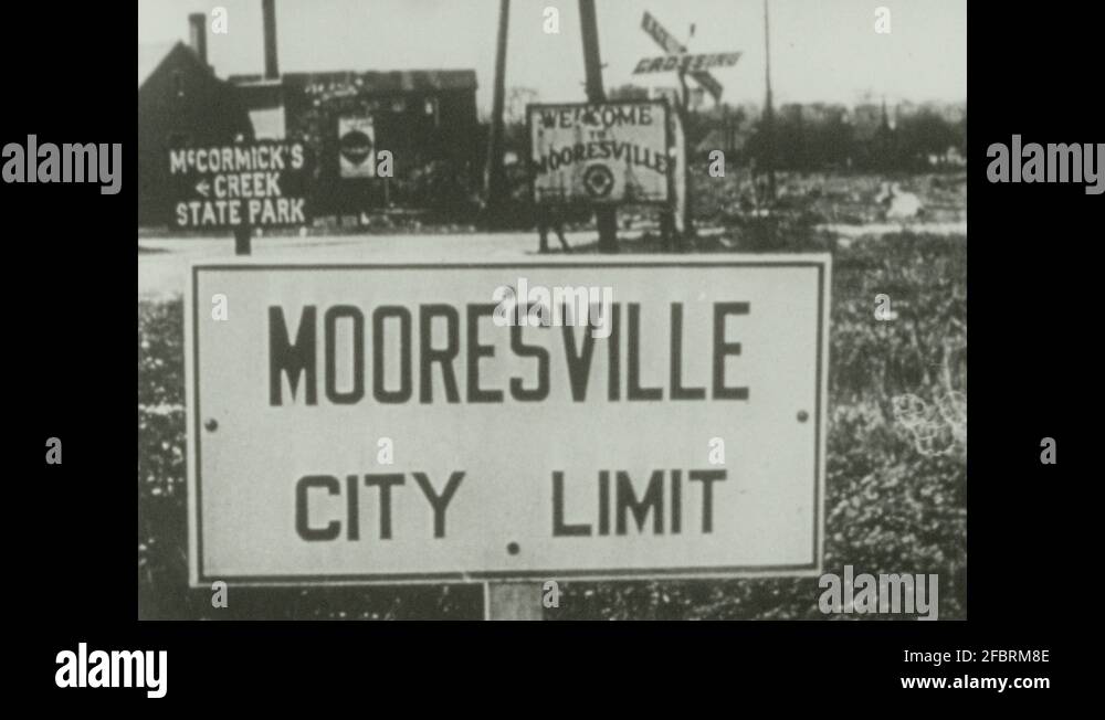 1930s: Sign for Mooresville. Street with buildings and cars. Cars drive ...