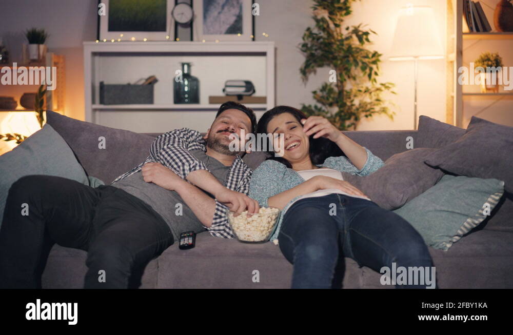 Happy people watching TV laughing eating popcorn lying on couch at home ...