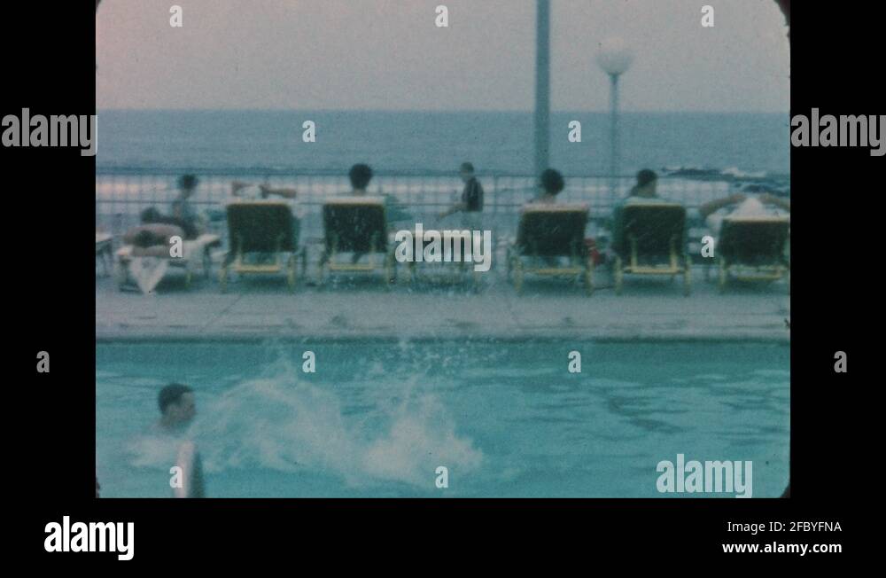 1960s: Girls dive from board into swimming pool. People lay and play on ...