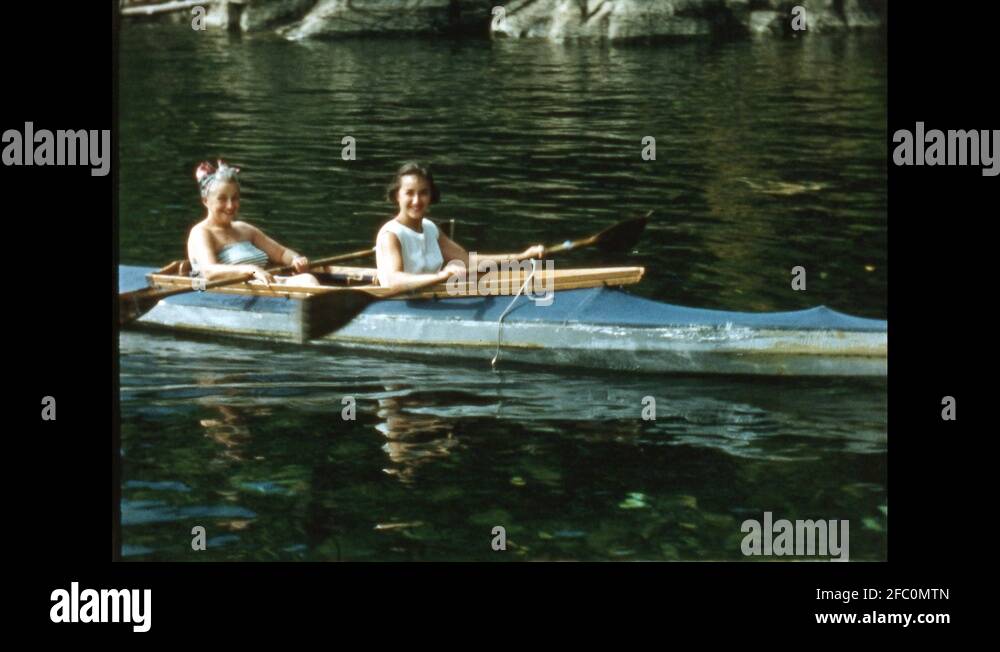 1960s: Women paddle canoe across lake. Girl in swimsuit walks on dock ...
