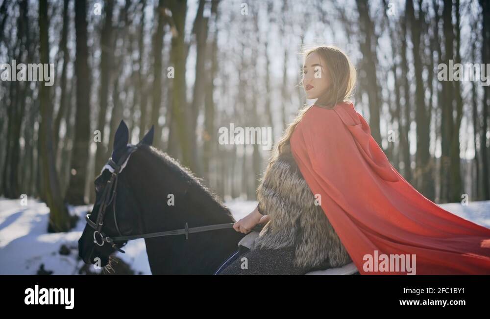 Woman in a red cloak riding a horse in winter during a snowfall in a ...