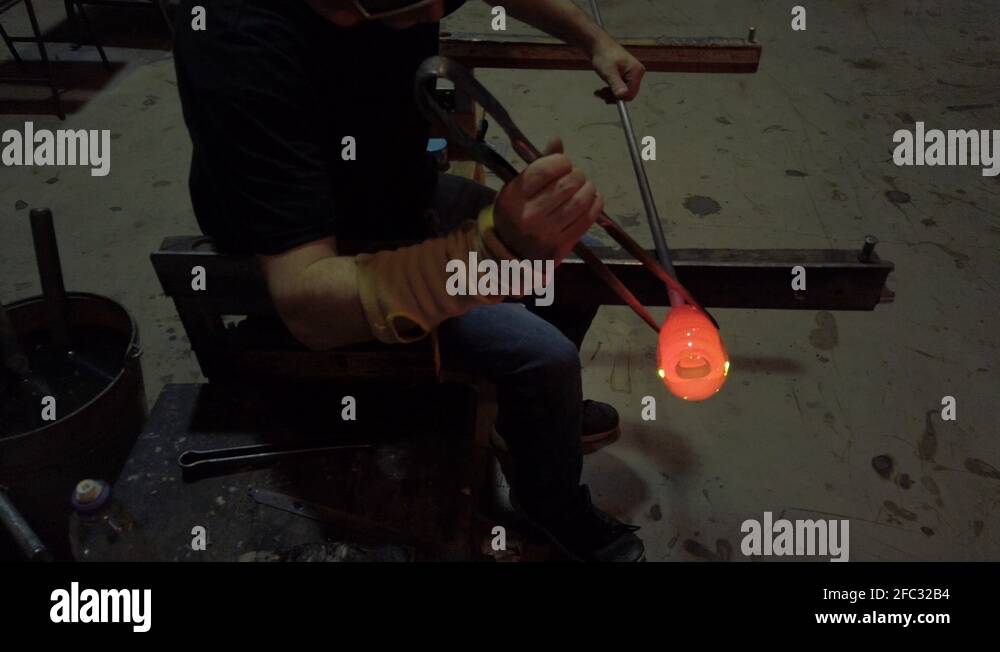 Craftsman shaping glowing hot glass into formed shape with metal tool ...