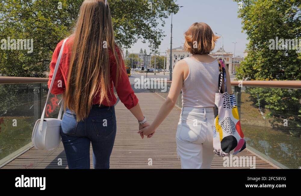 Two young women in love walk together in city holding hands back view ...