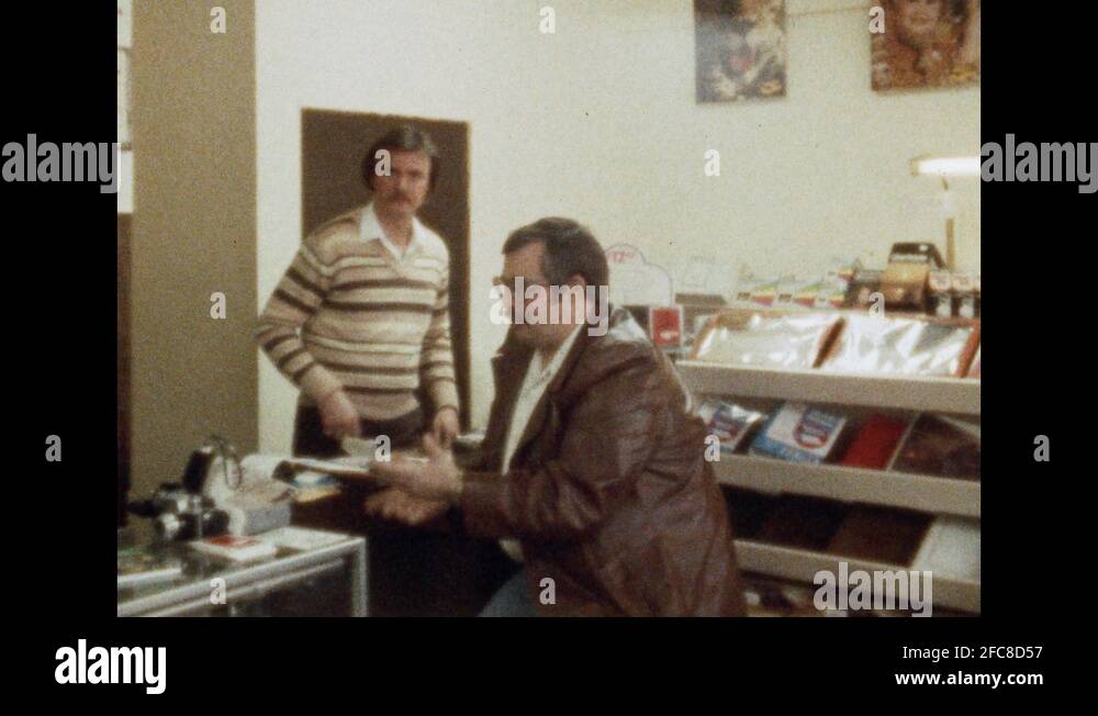 1970s: UNITED STATES: men work behind counter in shop. Men look at ...