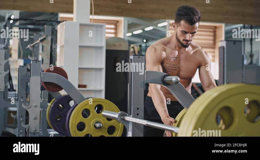 A handsome big muscular man is putting more weights into the bar , as ...