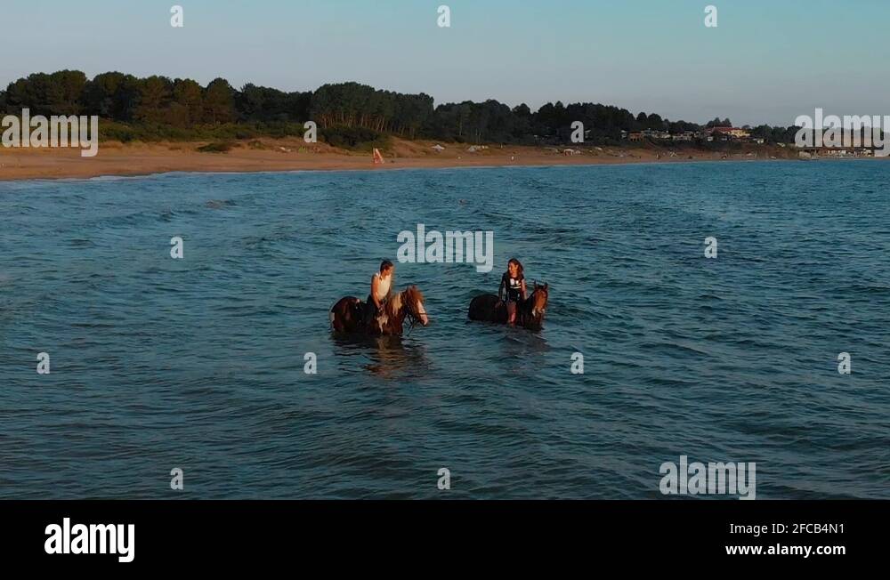 Two girls sit astride horses in shallow water laughing and being ...