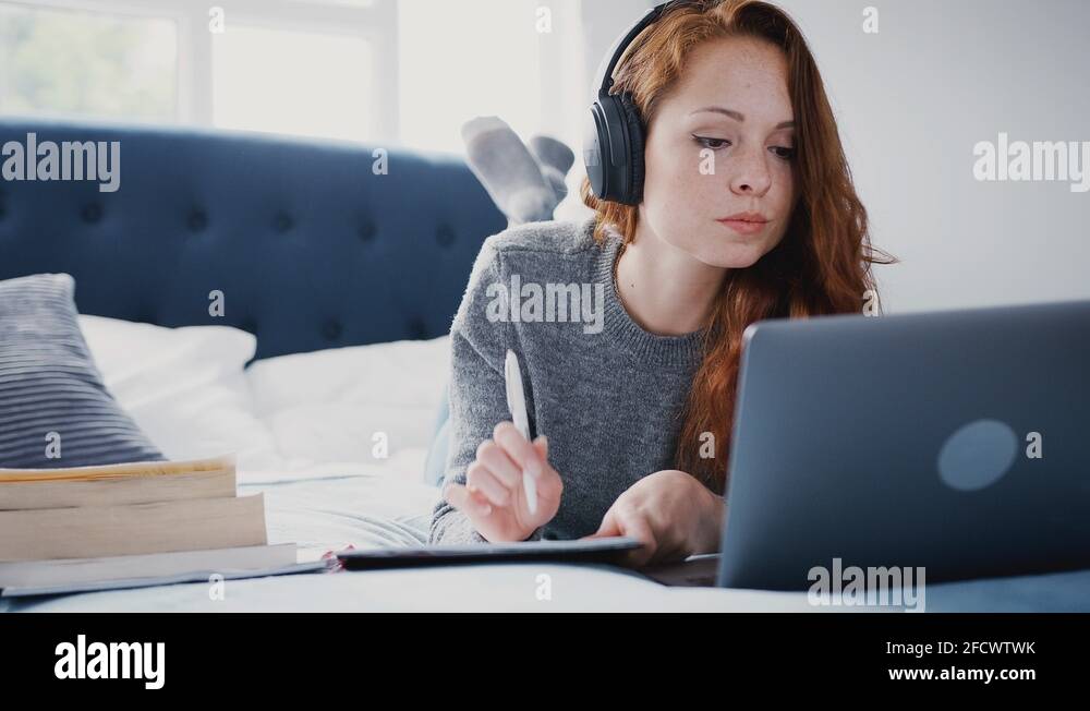 Female College Student Wearing Headphones Works On Bed In Shared House ...