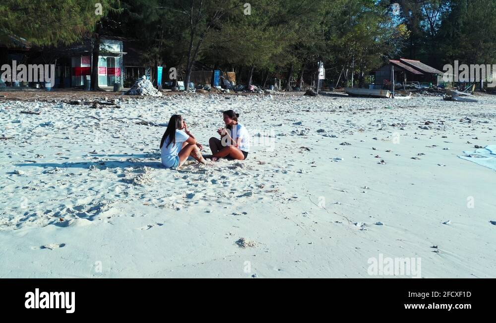 two teenage girls sitting on the white sand beach and laughing ...