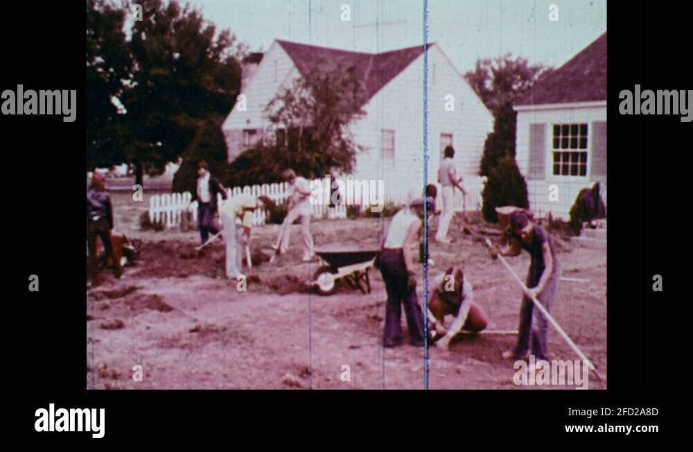 1970s: Large group of people rake and dig a front yard. Man clears the ...