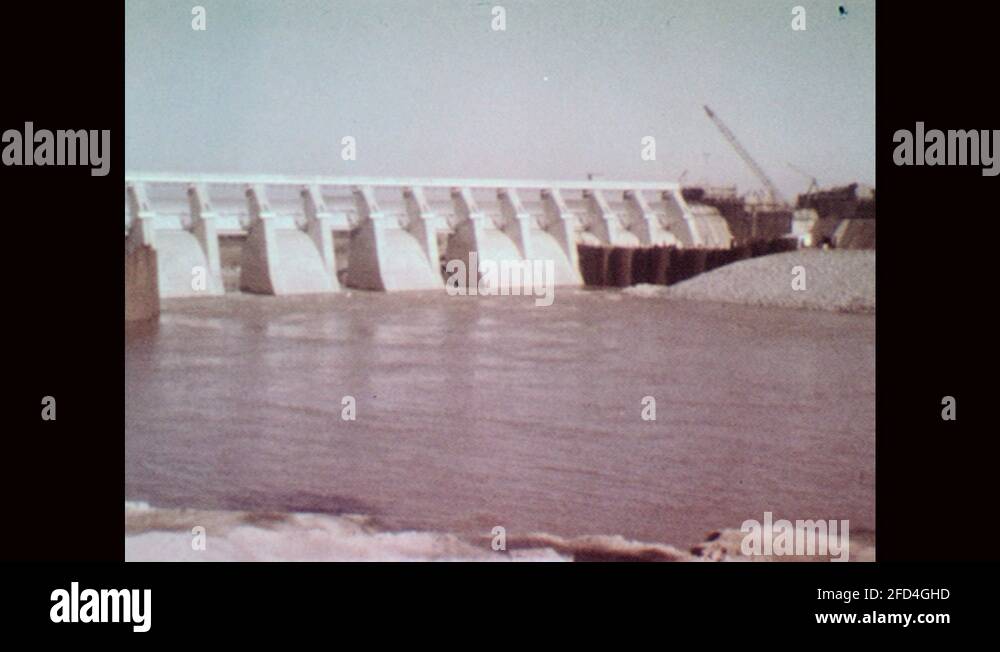 1960s: Water dam. Water tower. Man takes sample of water from shore ...