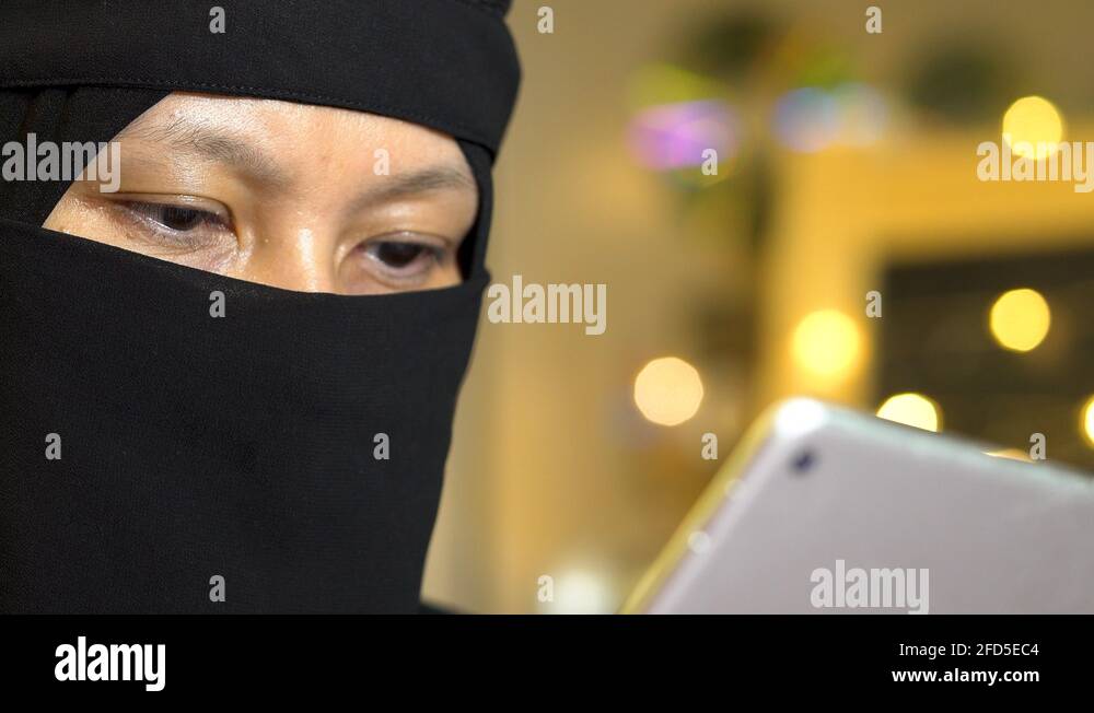 Niqab clad Muslim Asian Woman in Hijab alone reading on tablet close up ...