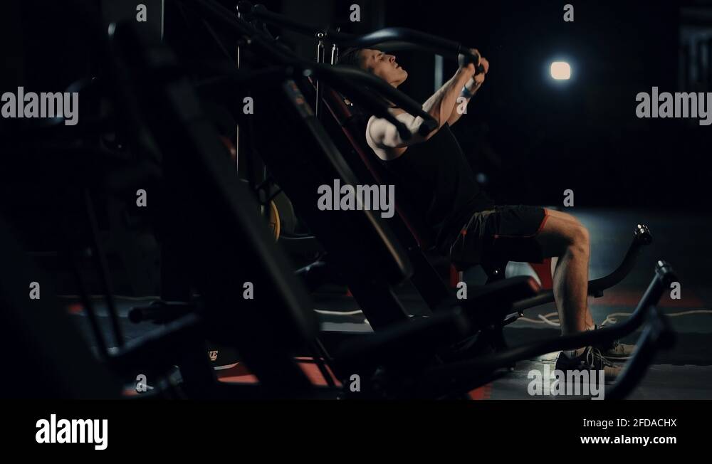 A muscular man performs exercises on pectoral muscles in a dark gym ...