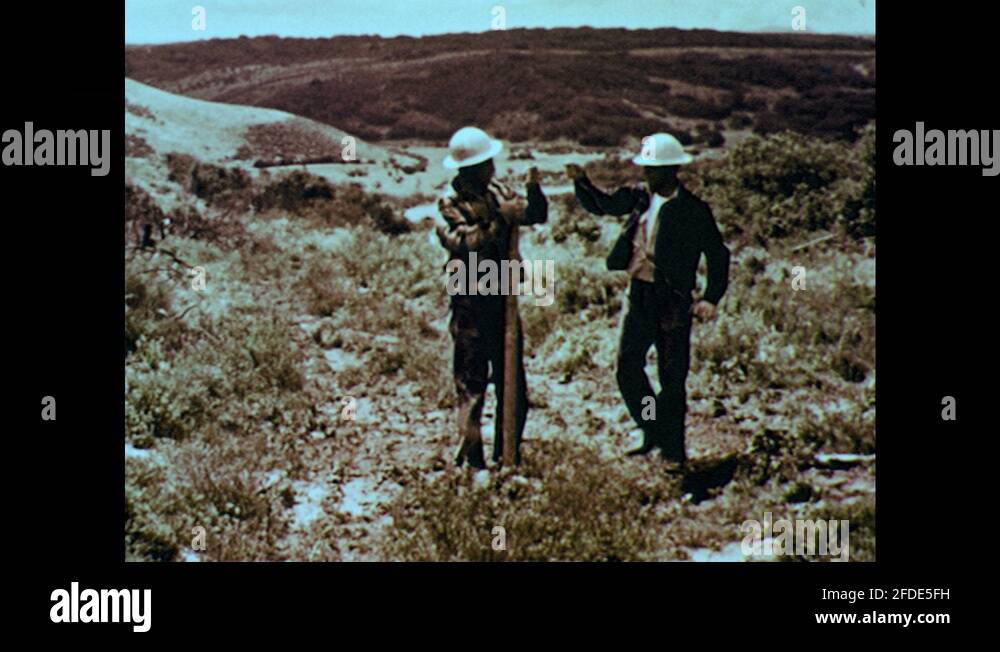 1950s: Two men lower large measuring stick into ground. Geyser of oil ...