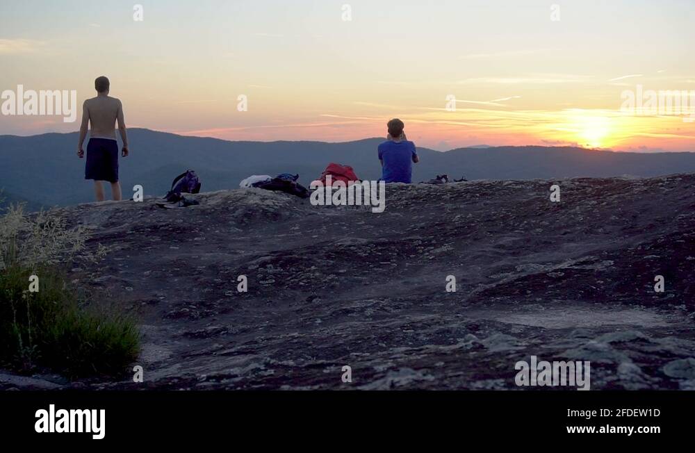two friends sitting on the edge of a cliff and watching a sunset ...