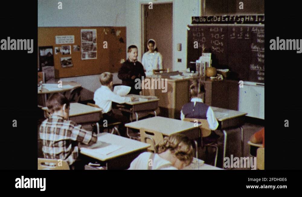 1960s: children walk into classroom, teacher bends to help girl take ...