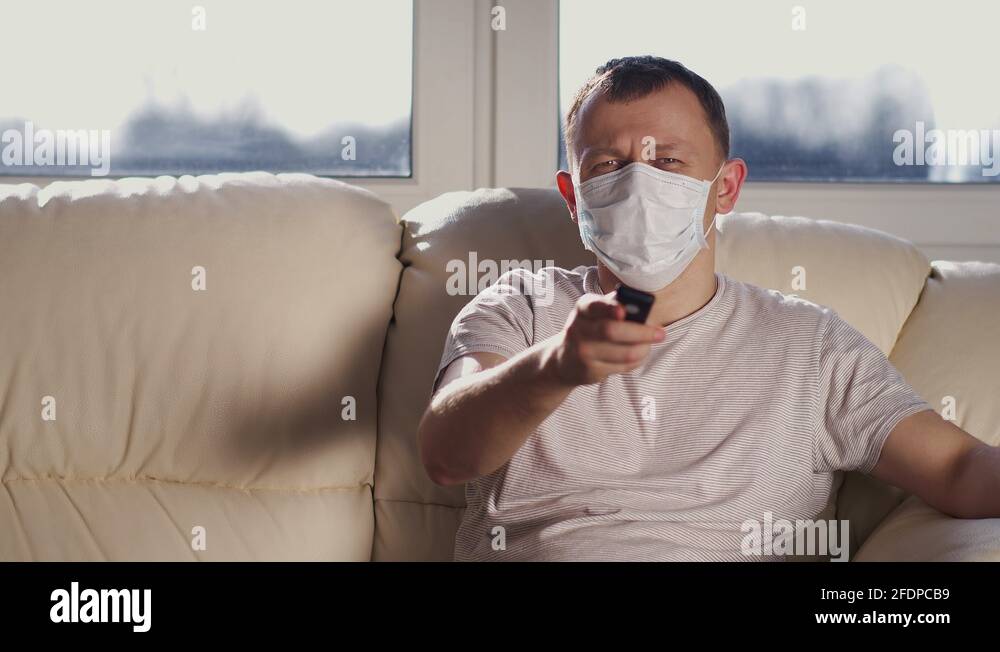 A man in a face mask sits at home on the couch and switches the TV ...