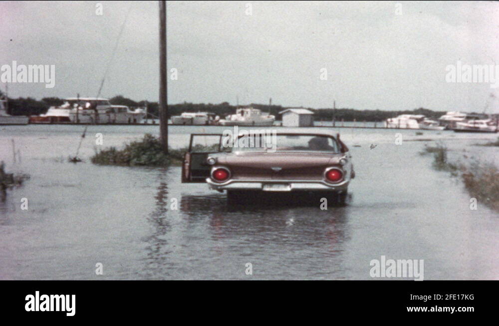1960s: People sit in car with door open in flooded water. Storm blows ...