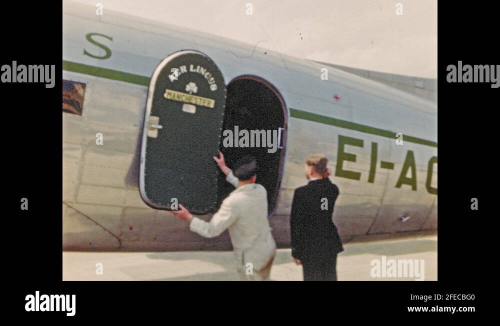 1950s: Stewardess and porter stand near stairs on runway. Men pull ...