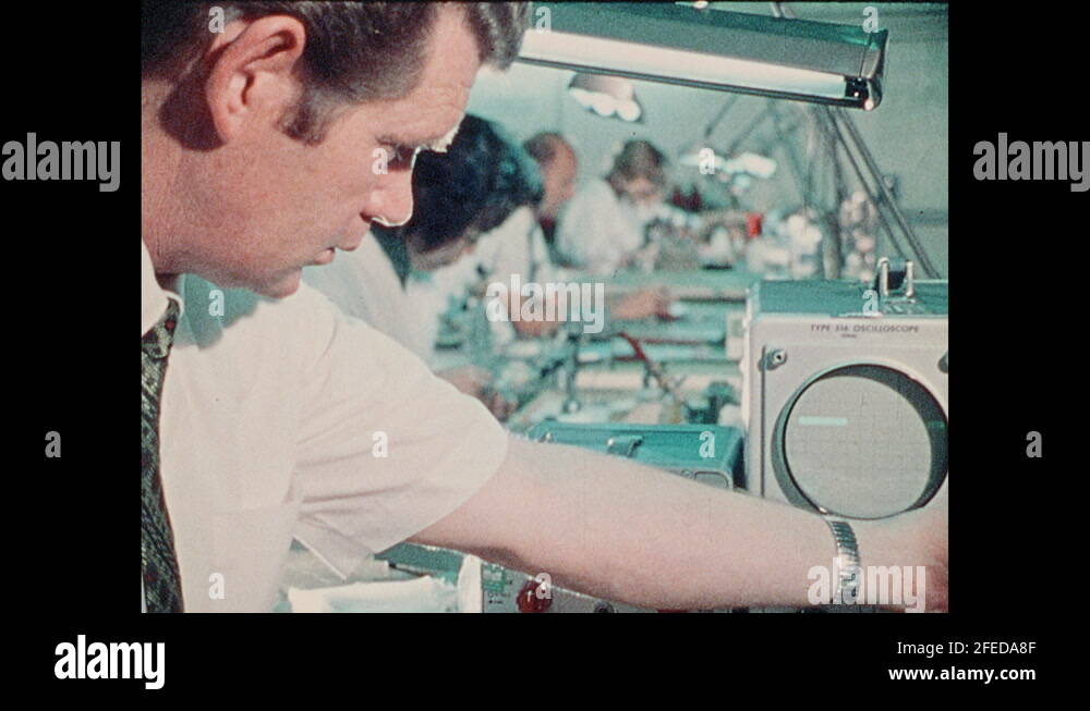 1970s: People at work in lab. Woman uses soldering iron. Man marks ...
